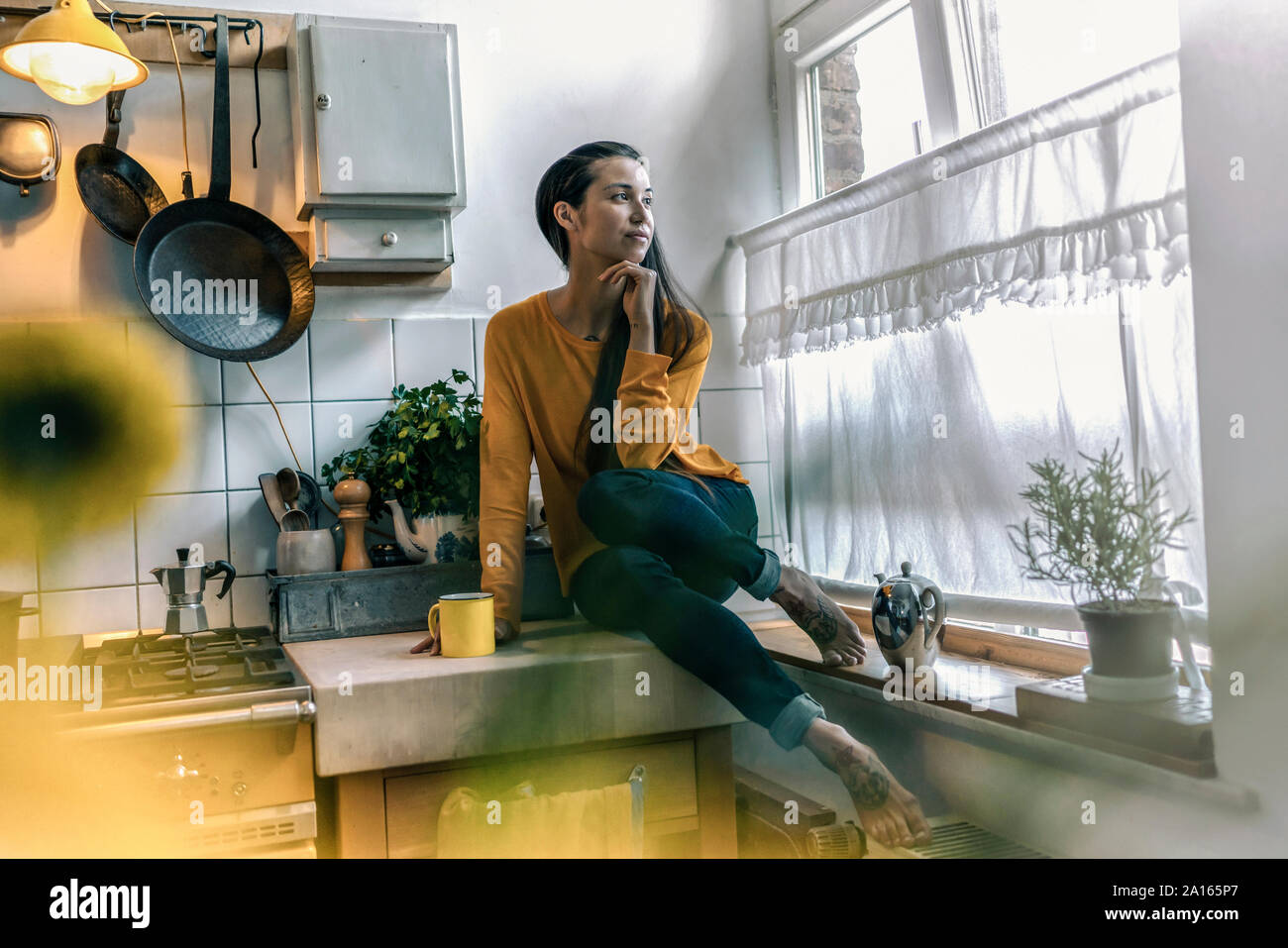 Young woman sitting on kitchen counter at home looking out of window ...