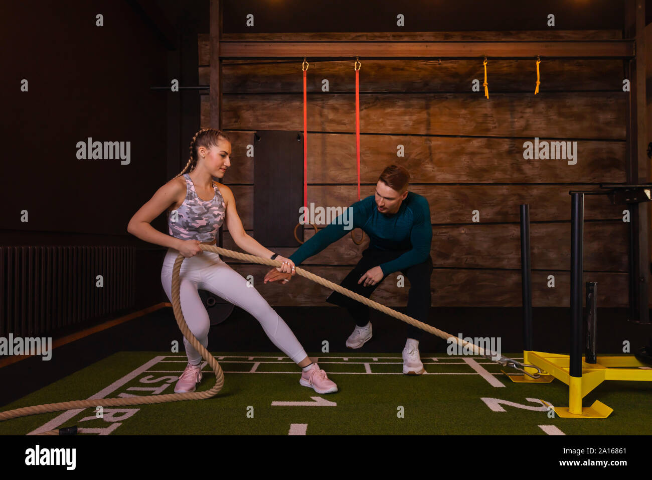 Young woman exercising in fitness gym with personal trainer pulling ...