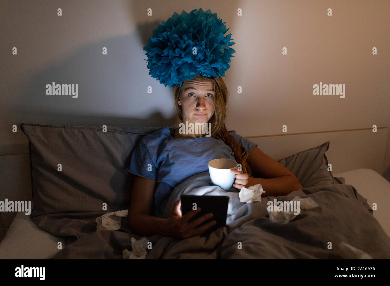 Ill young woman lying in bed at home with cup of tea at night using ...