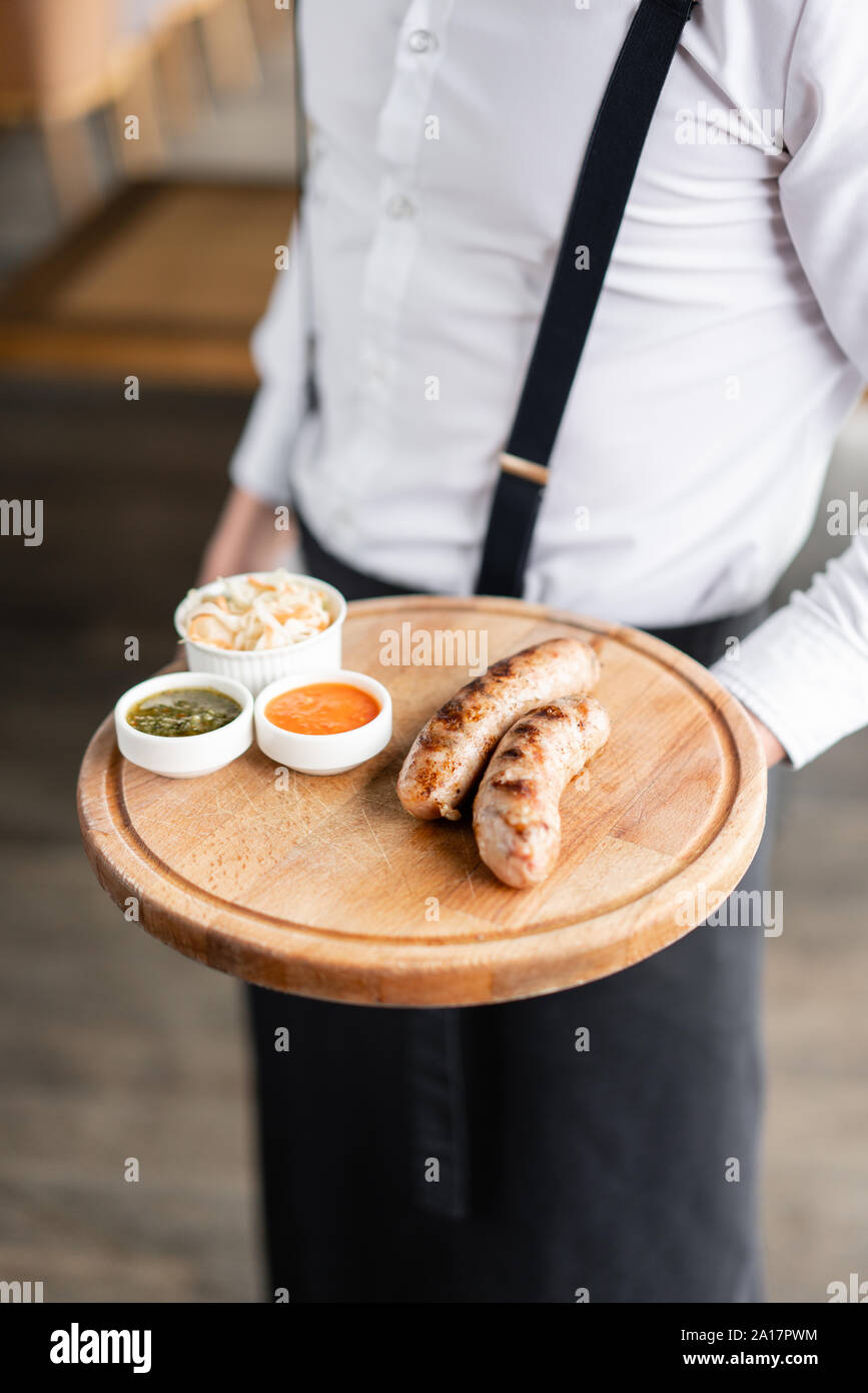 The waiter is holding plate with Grilled sausages. Serving on a wooden ...