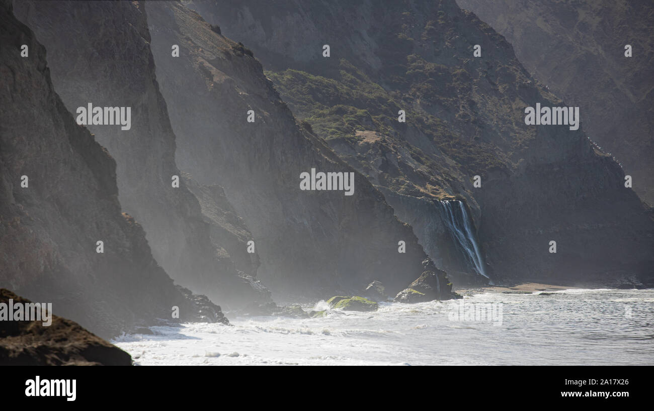 Waterfall flows into sea amid steep cliffs and rugged coastal landscape ...