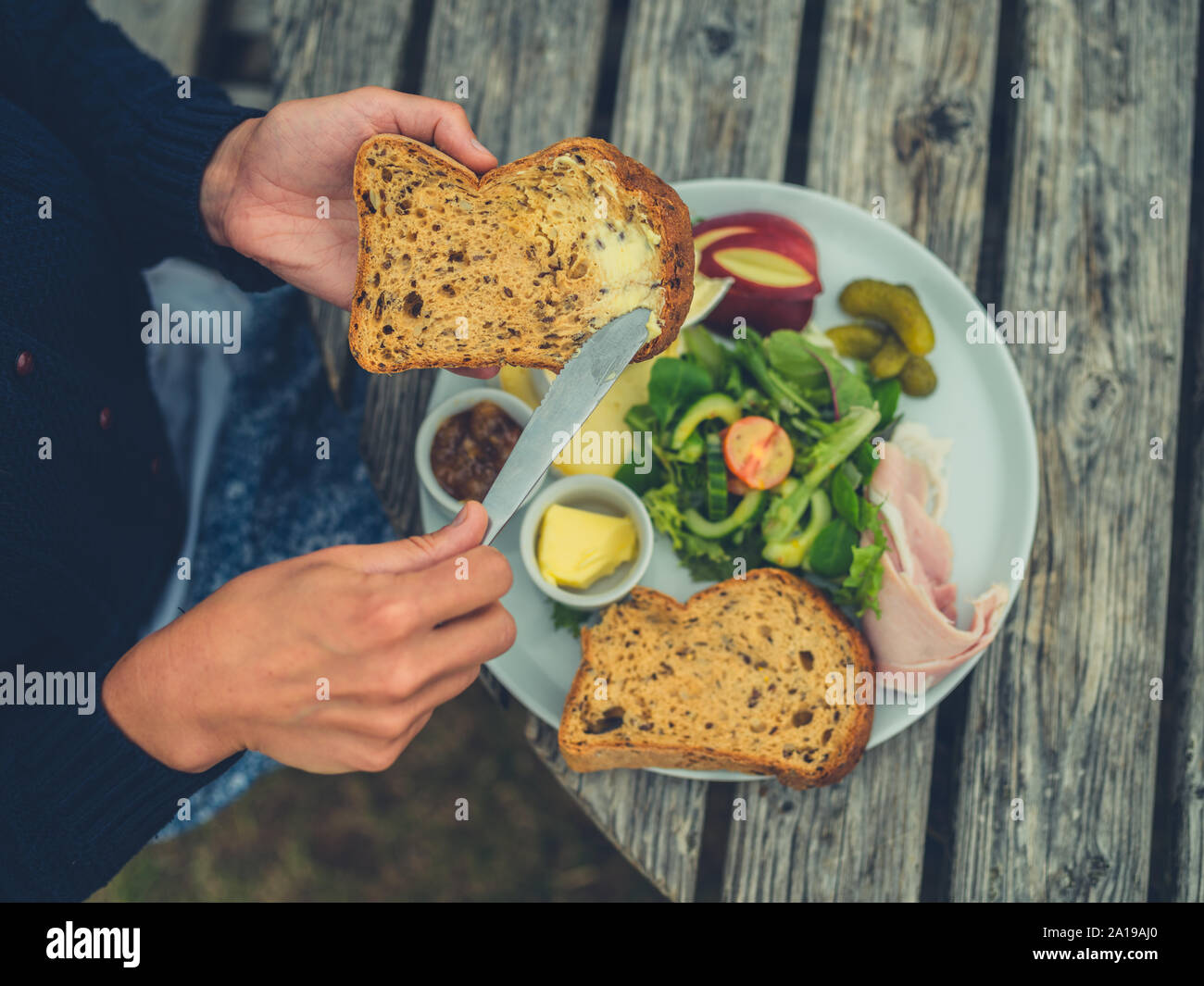 A young woman is buttering bread outdoors at a picnic table to make a ...