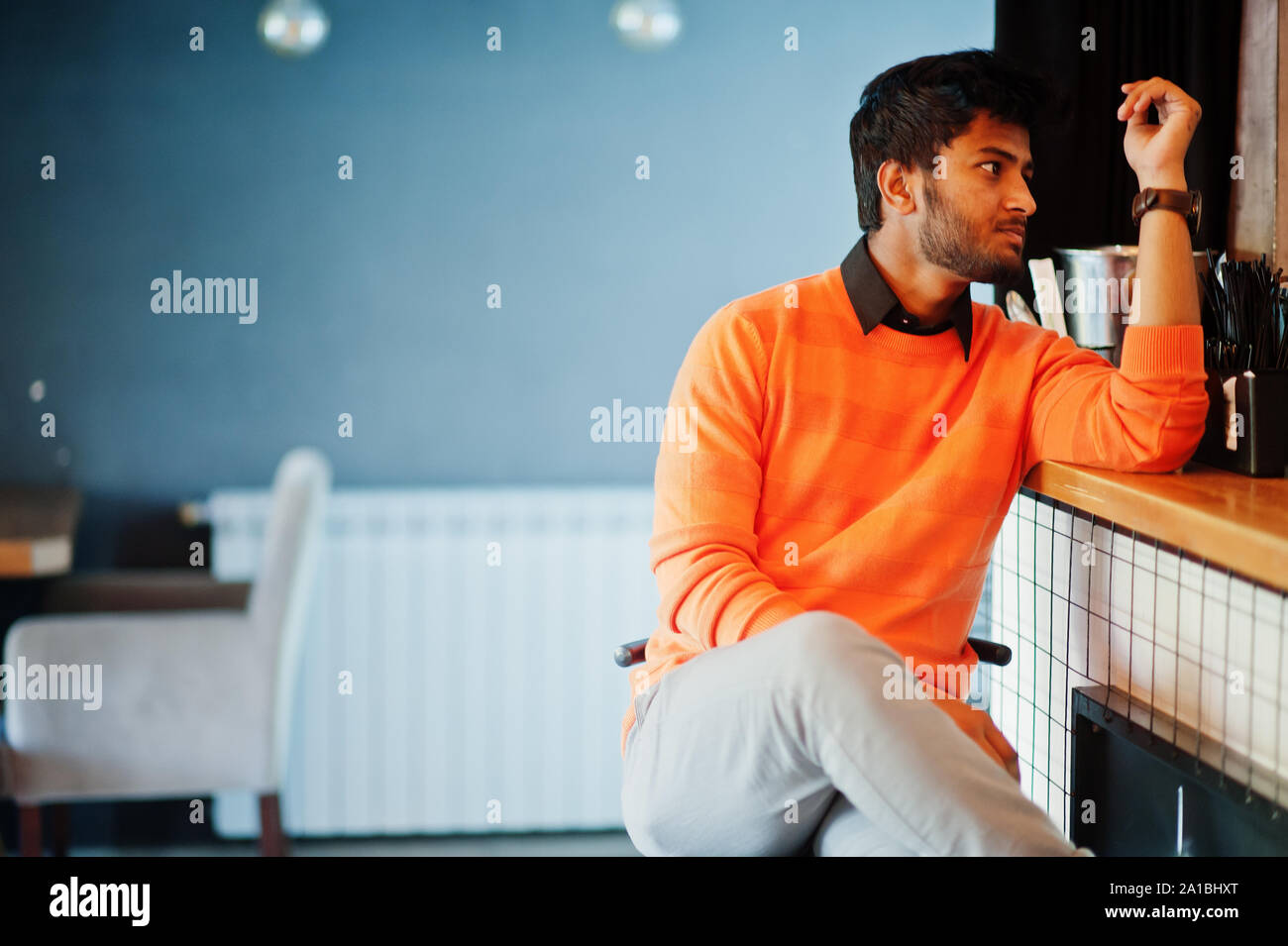Confident young indian man in orange sweater sitting bar counter at ...