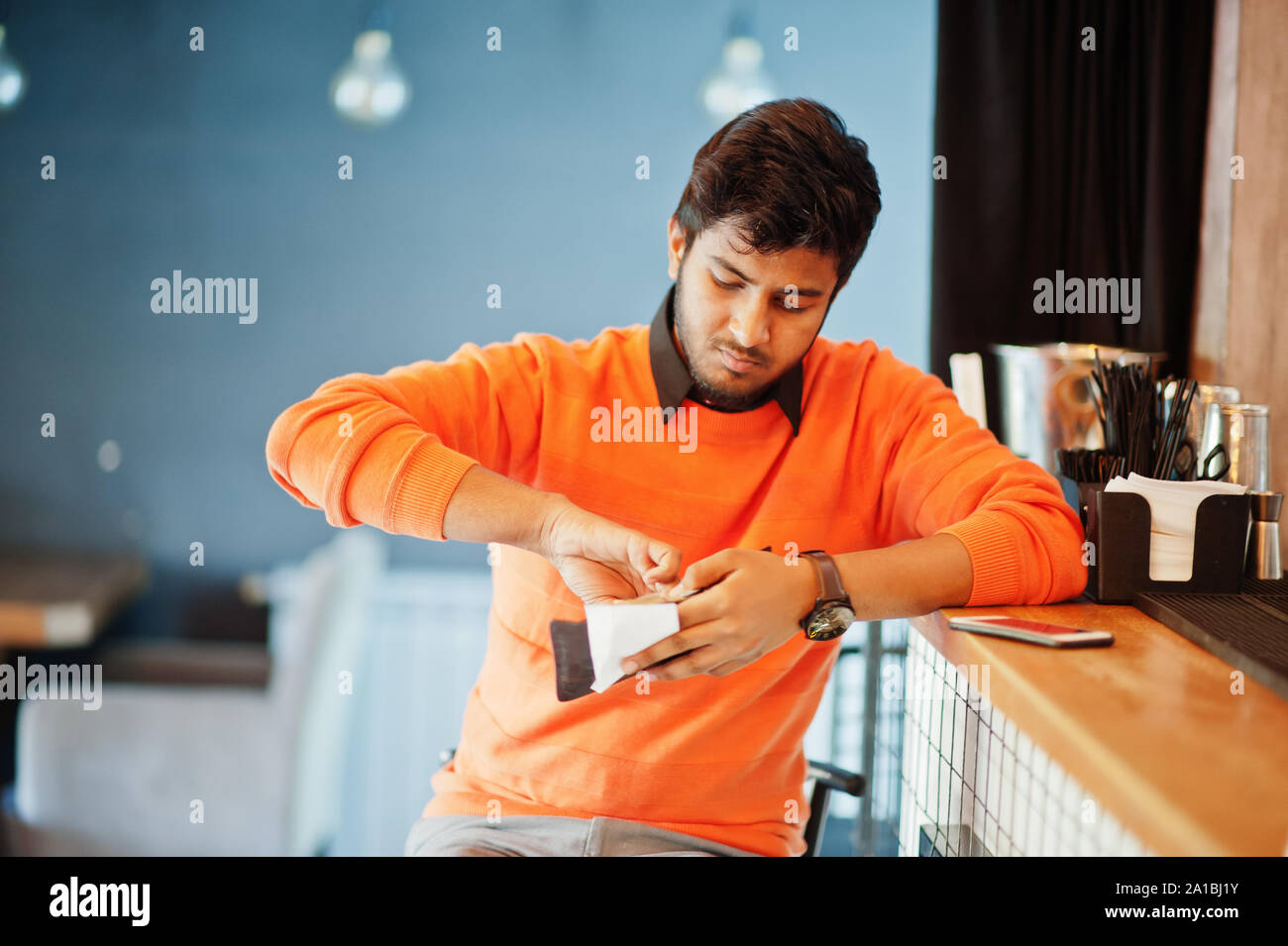 Confident young indian man in orange sweater sitting bar counter at ...