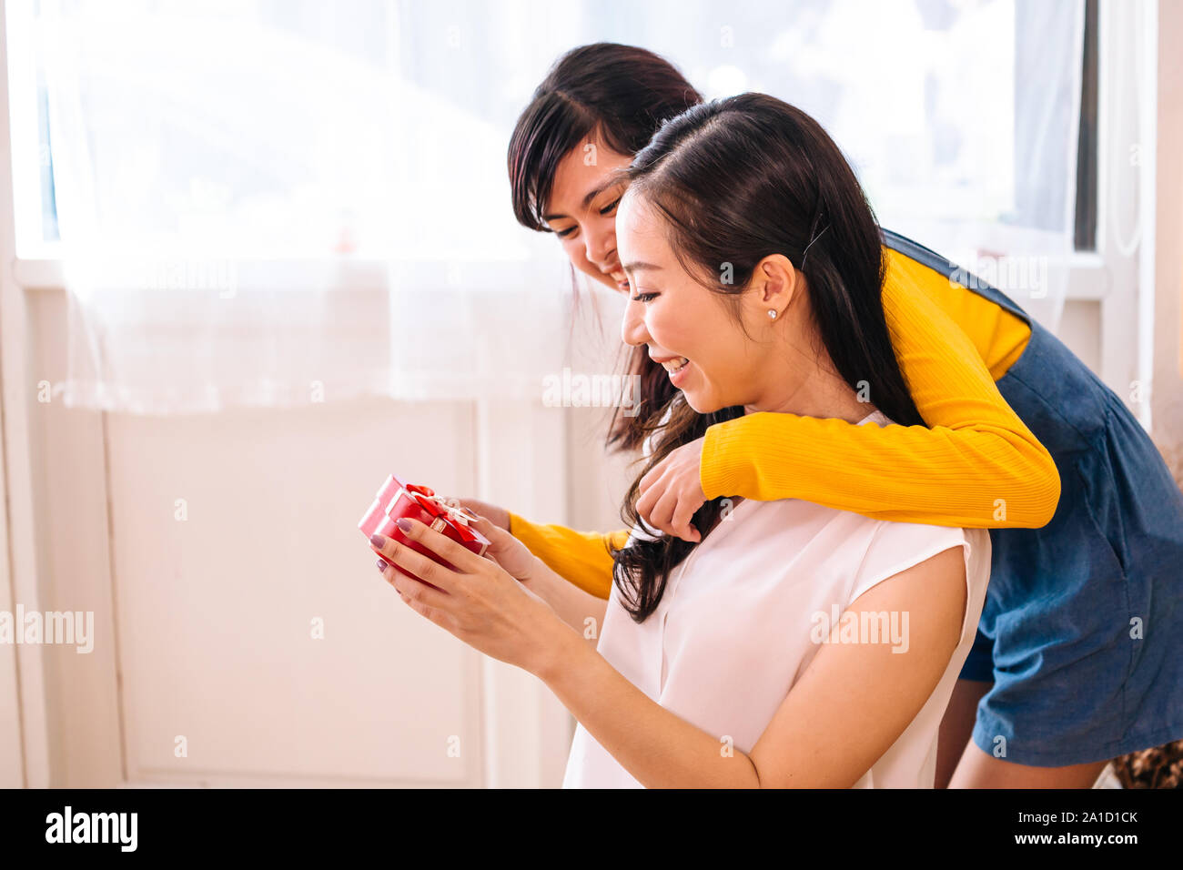 Smiling happy Asian teenage daughter and Asian middle-aged mother hugging together in indoor ...
