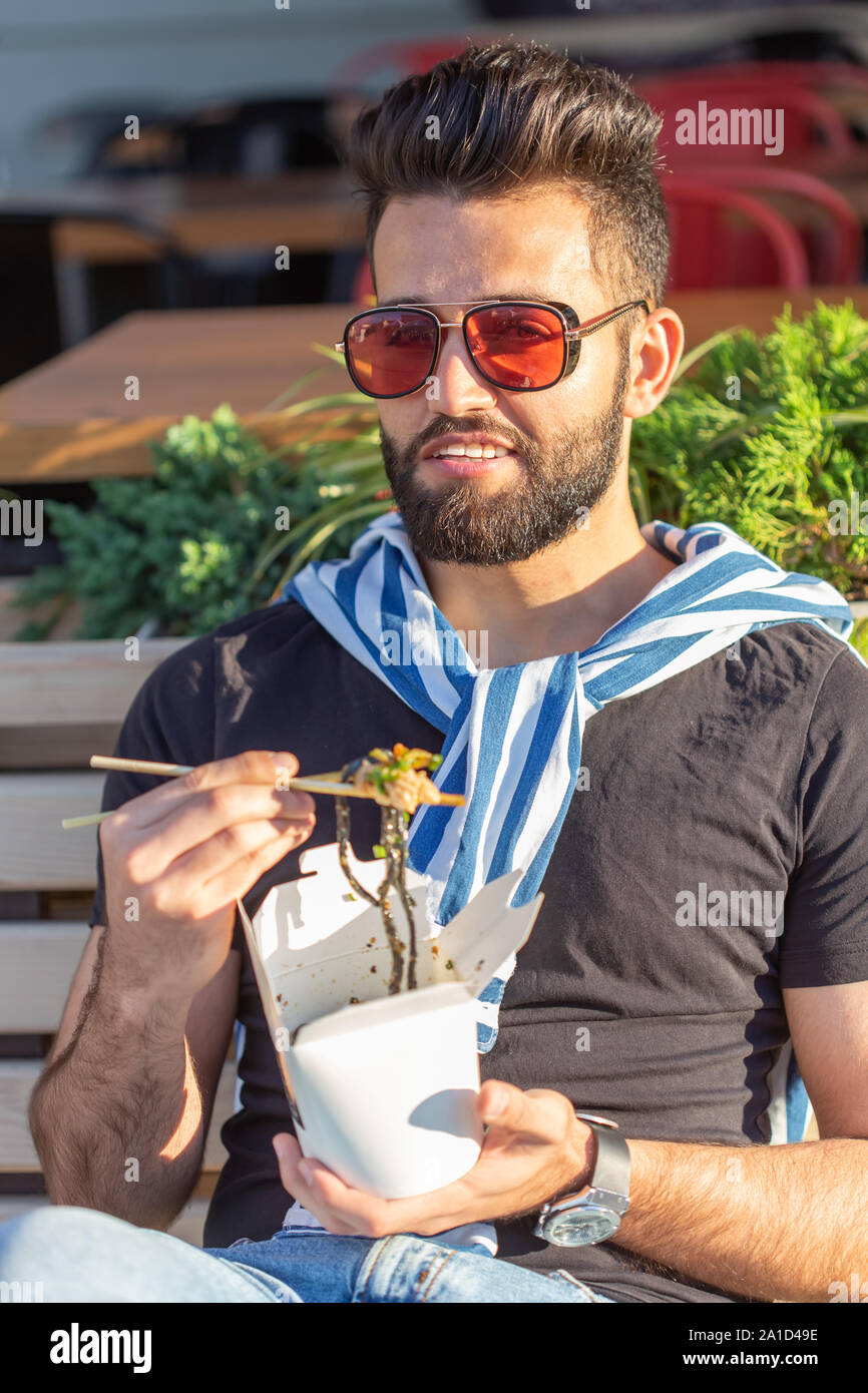 Young male hipster with a mustache and beard is eating chinese noodles ...