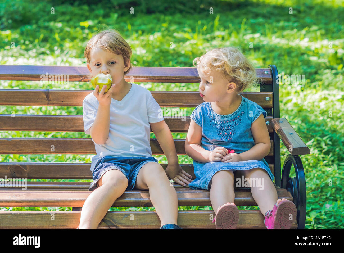 Toddlers boy and girl sitting on a bench by the sea and eat an apple ...