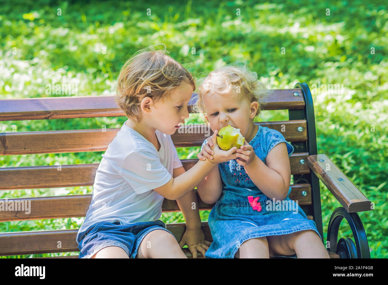 Toddlers boy and girl sitting on a bench by the sea and eat an apple ...
