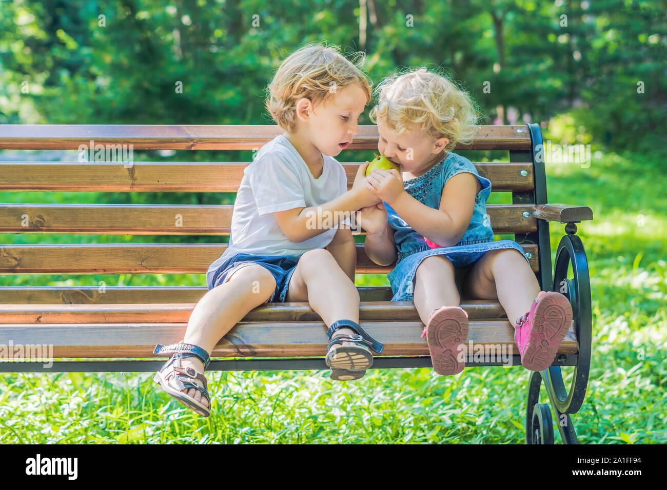 Toddlers boy and girl sitting on a bench by the sea and eat an apple ...