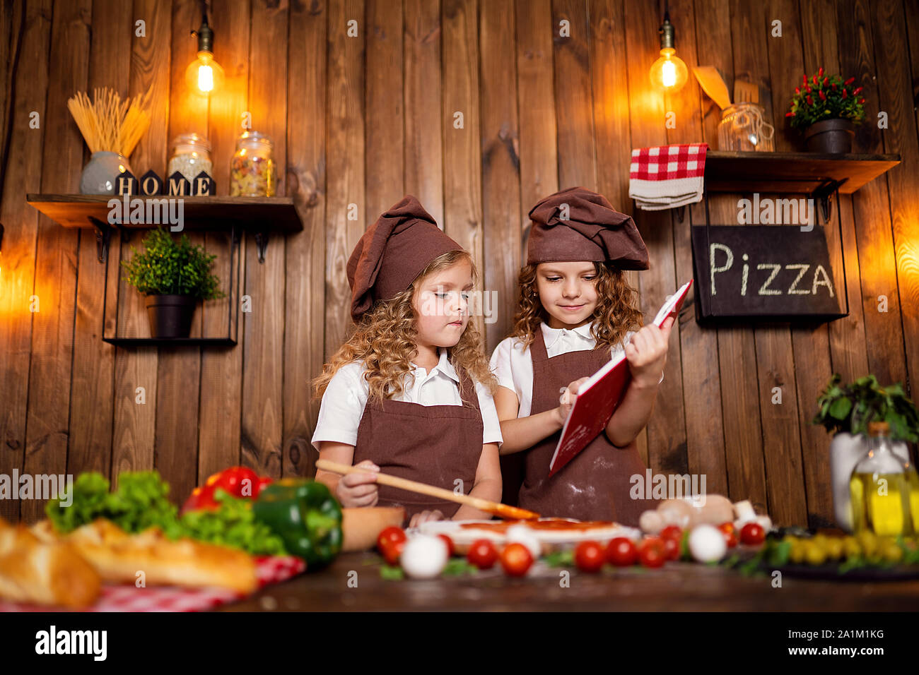 Cute girls in aprons and hats reading recipe book together while ...