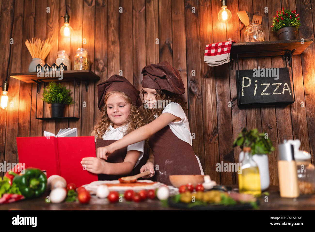 Cute girls in aprons and hats reading recipe book together while ...