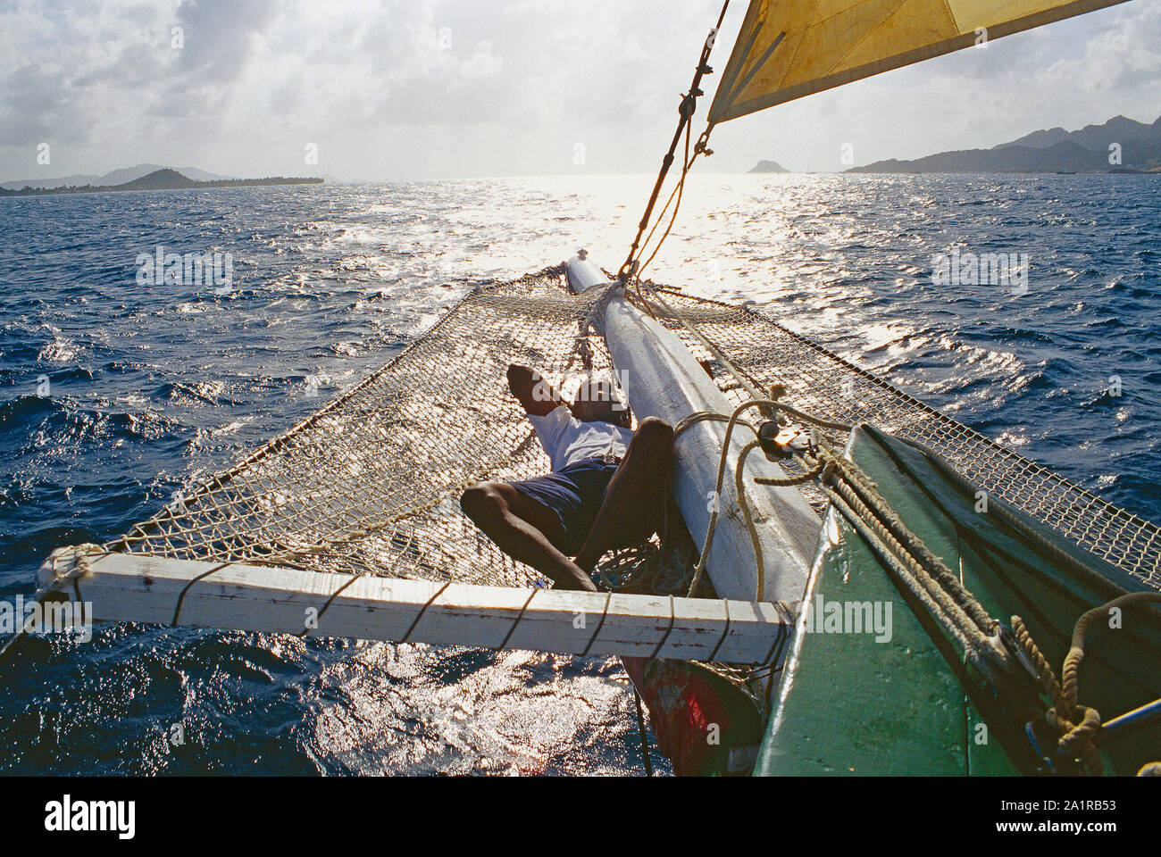 Barbados. Local man relaxing on bowsprit safety net of sailing yacht ...