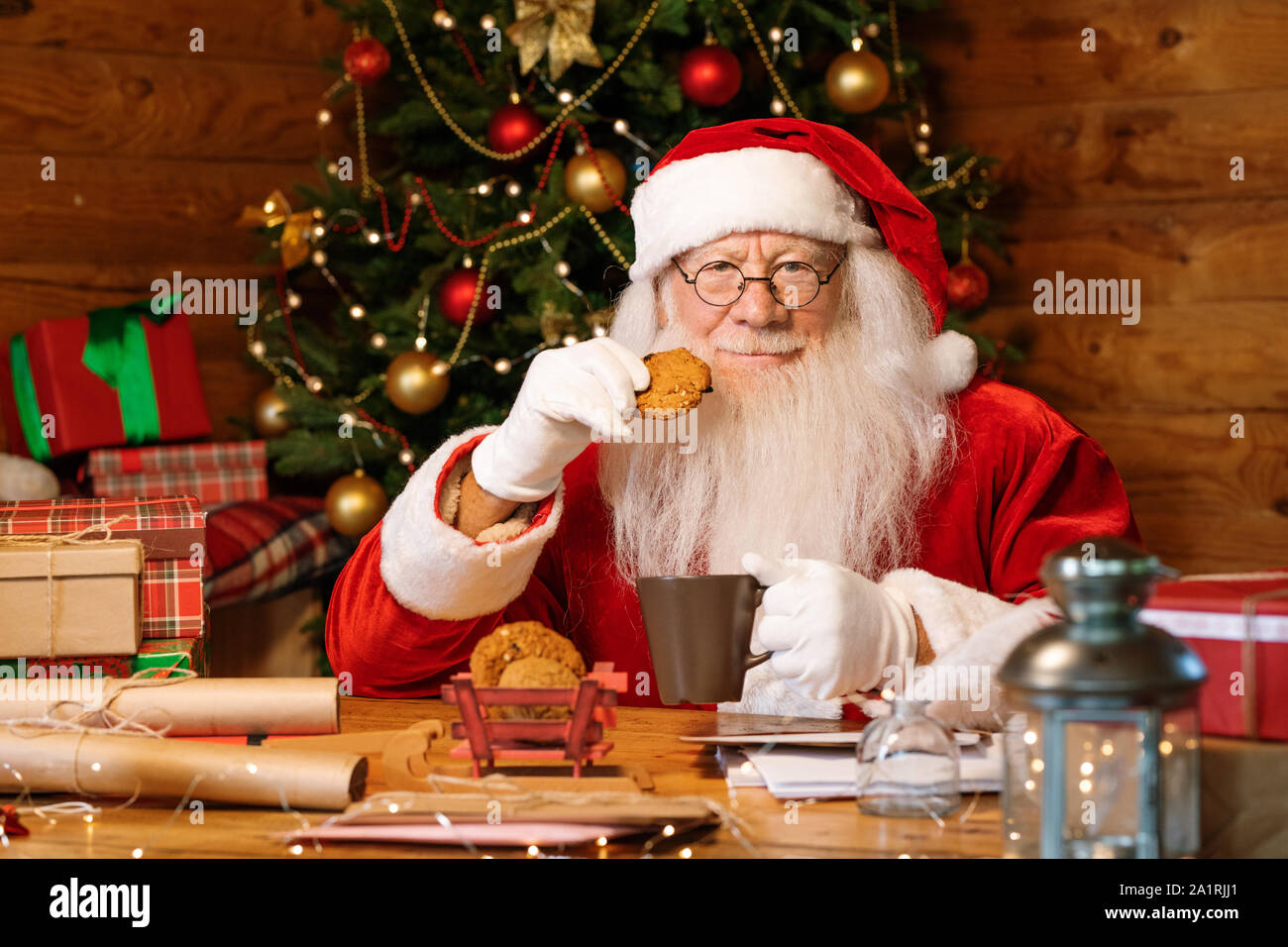 Hungry Santa in costume sitting by table and eating cookies with milk ...