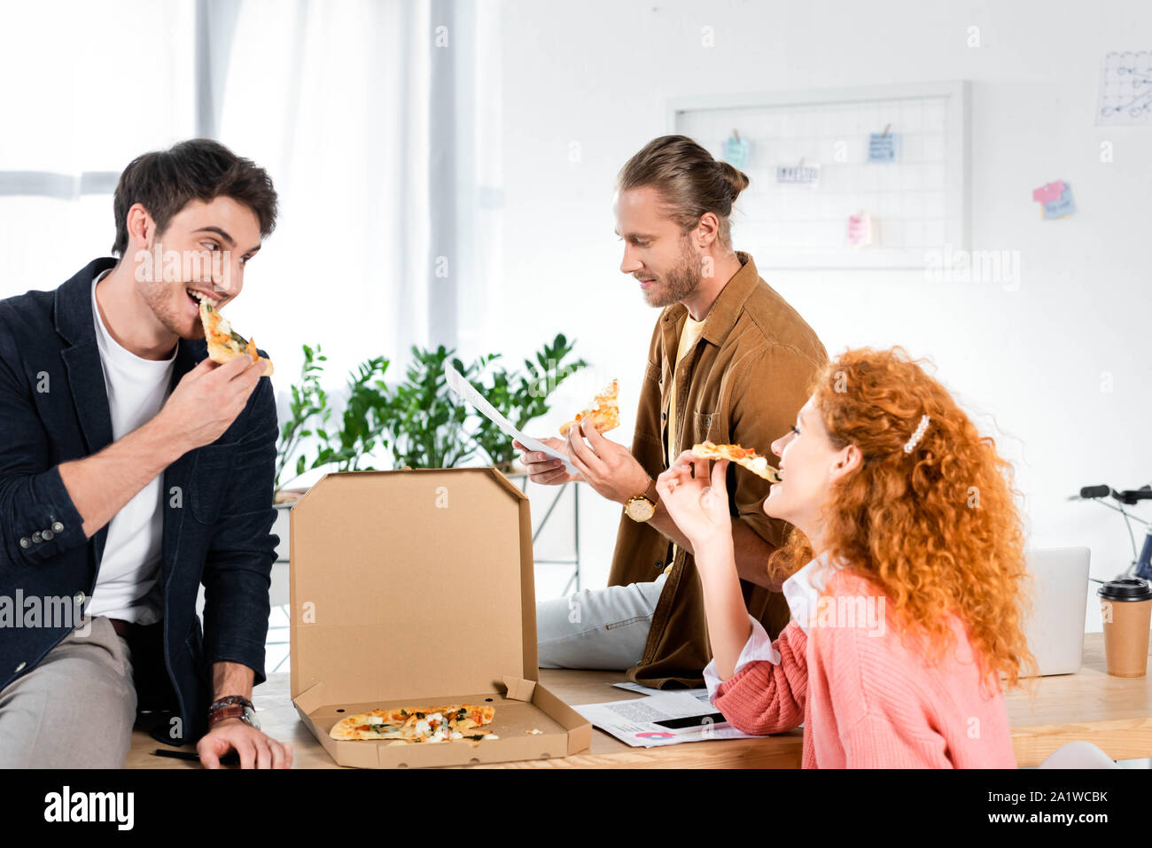 three smiling friends eating pizza and doing paperwork in office Stock ...