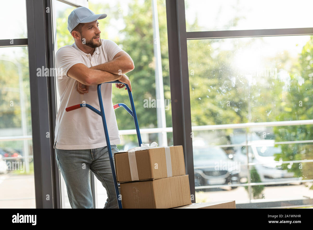 handsome delivery man standing near delivery cart with cardboard boxes ...