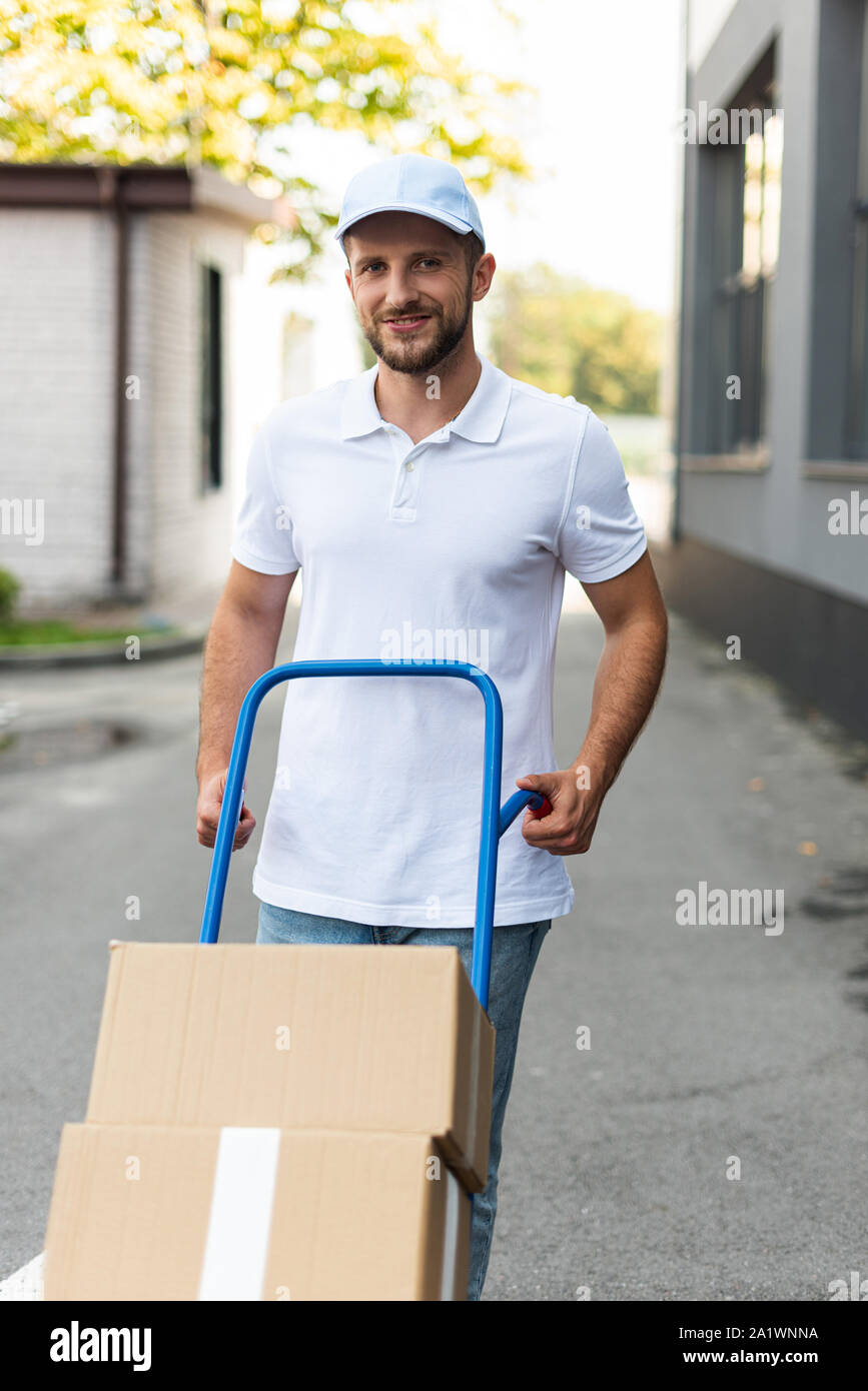 positive delivery man standing with delivery cart near building Stock ...