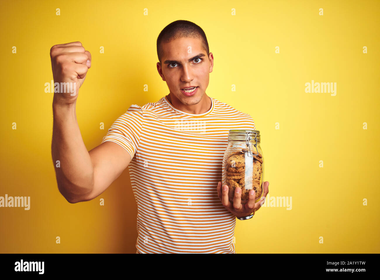 Young handsome man holding a jar of cookies over yellow isolated ...