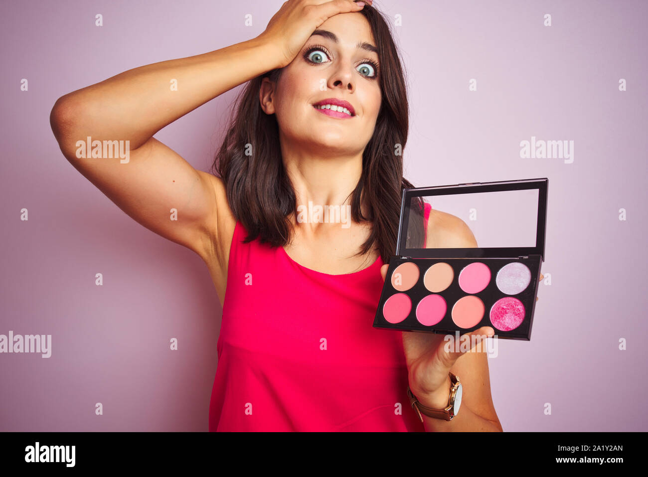 Young beautiful woman applying make up over pink isolated background ...