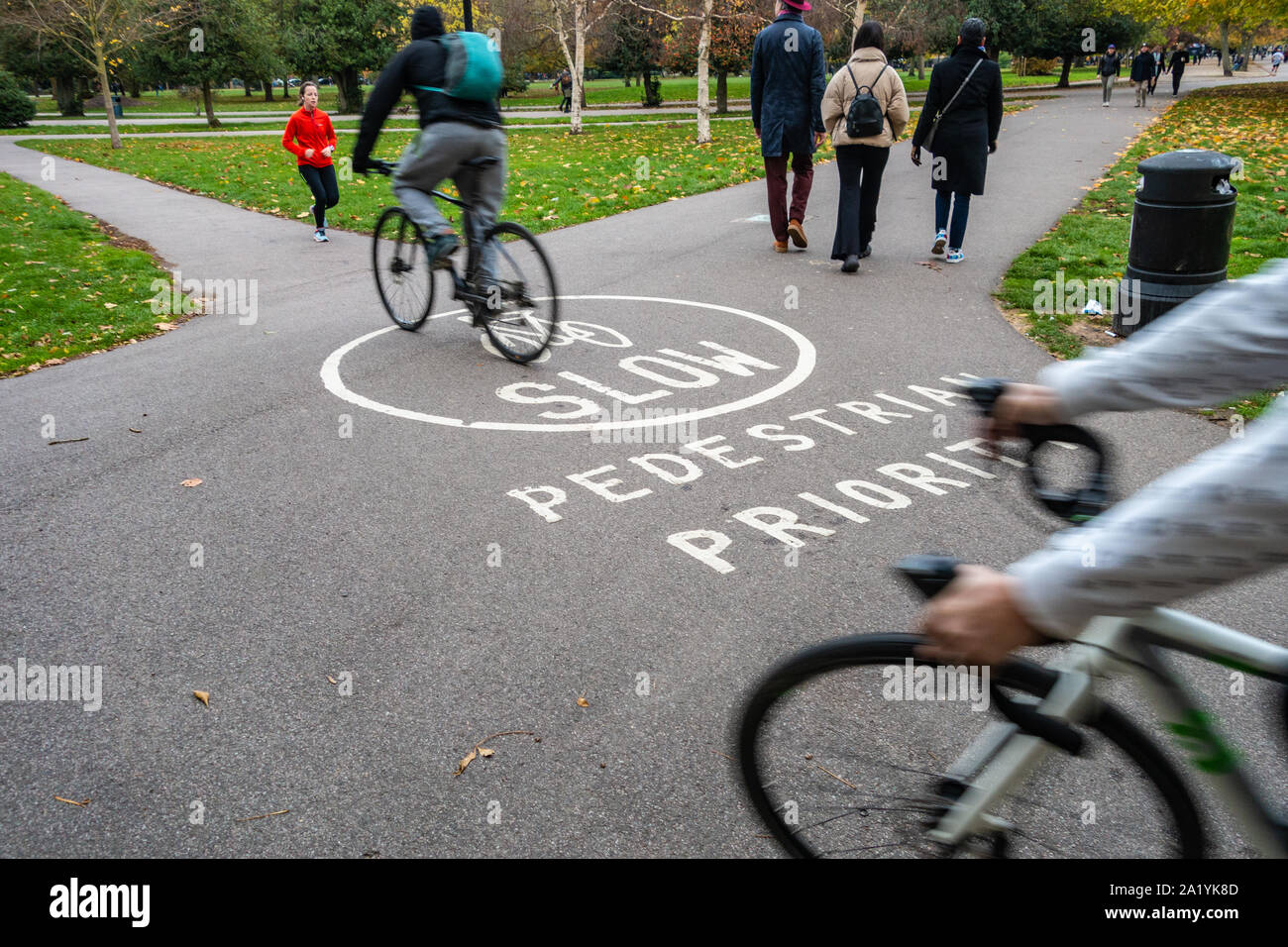 Cyclists riding over a pedestrian priority warning sign on a park ...