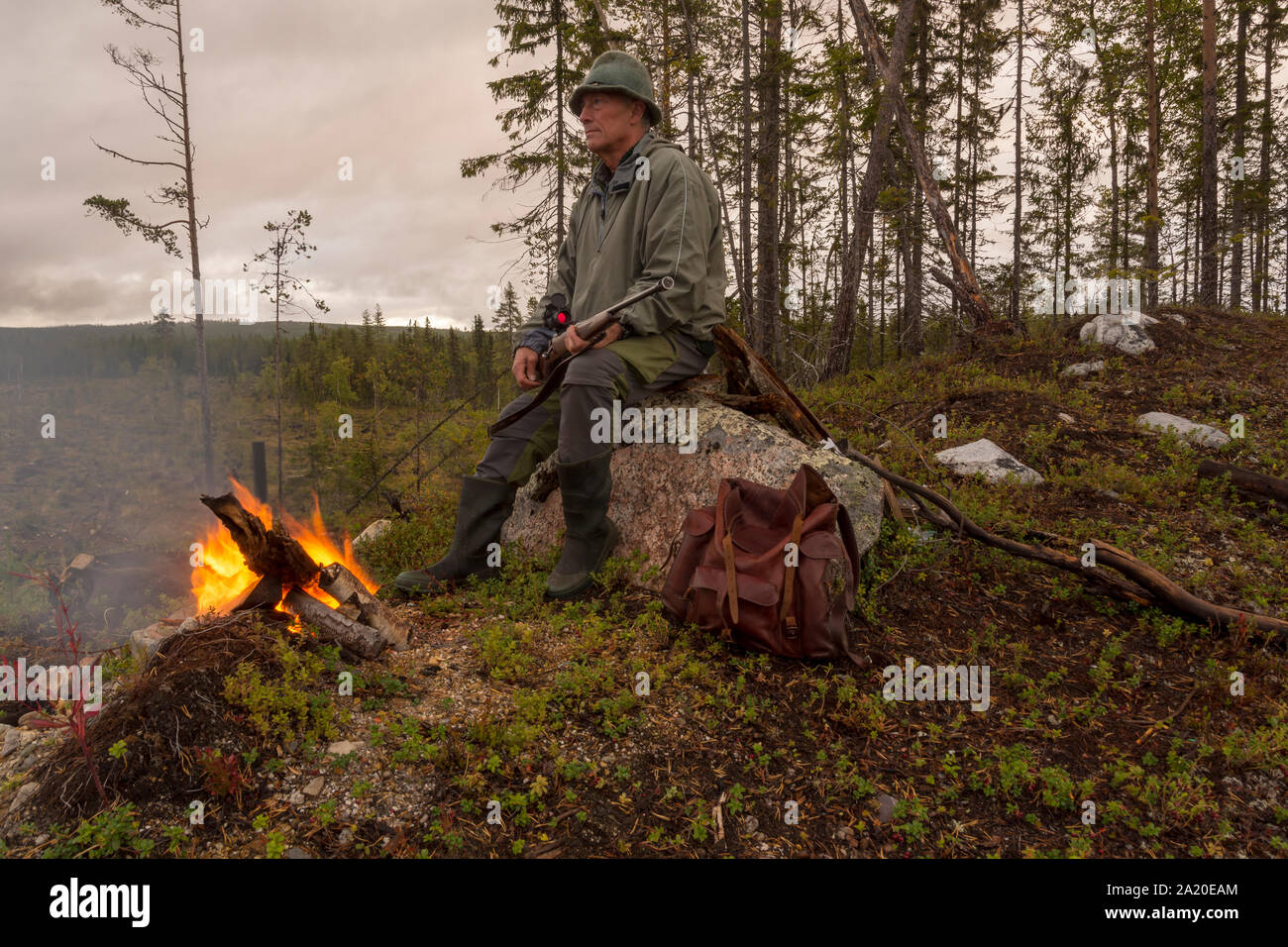 Moose hunter sitting on a rock holding his rifle with a campfire in ...