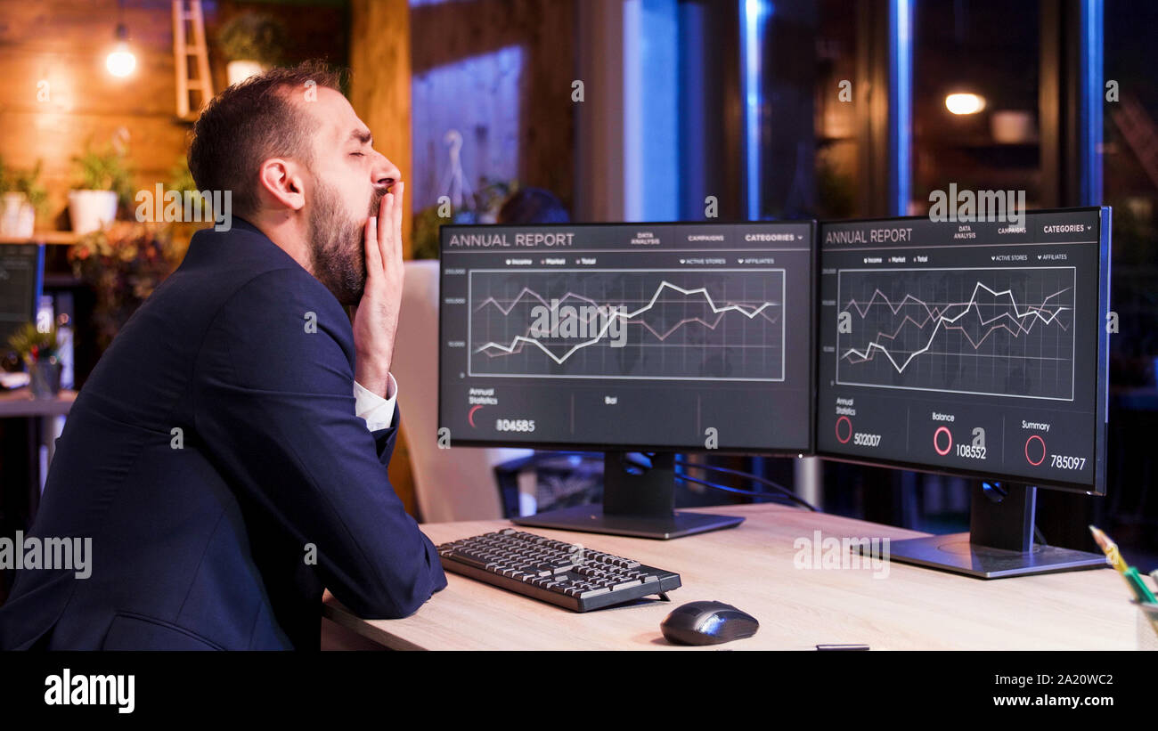 Overworked businessman yawning at his office desk late at night. Blue ...