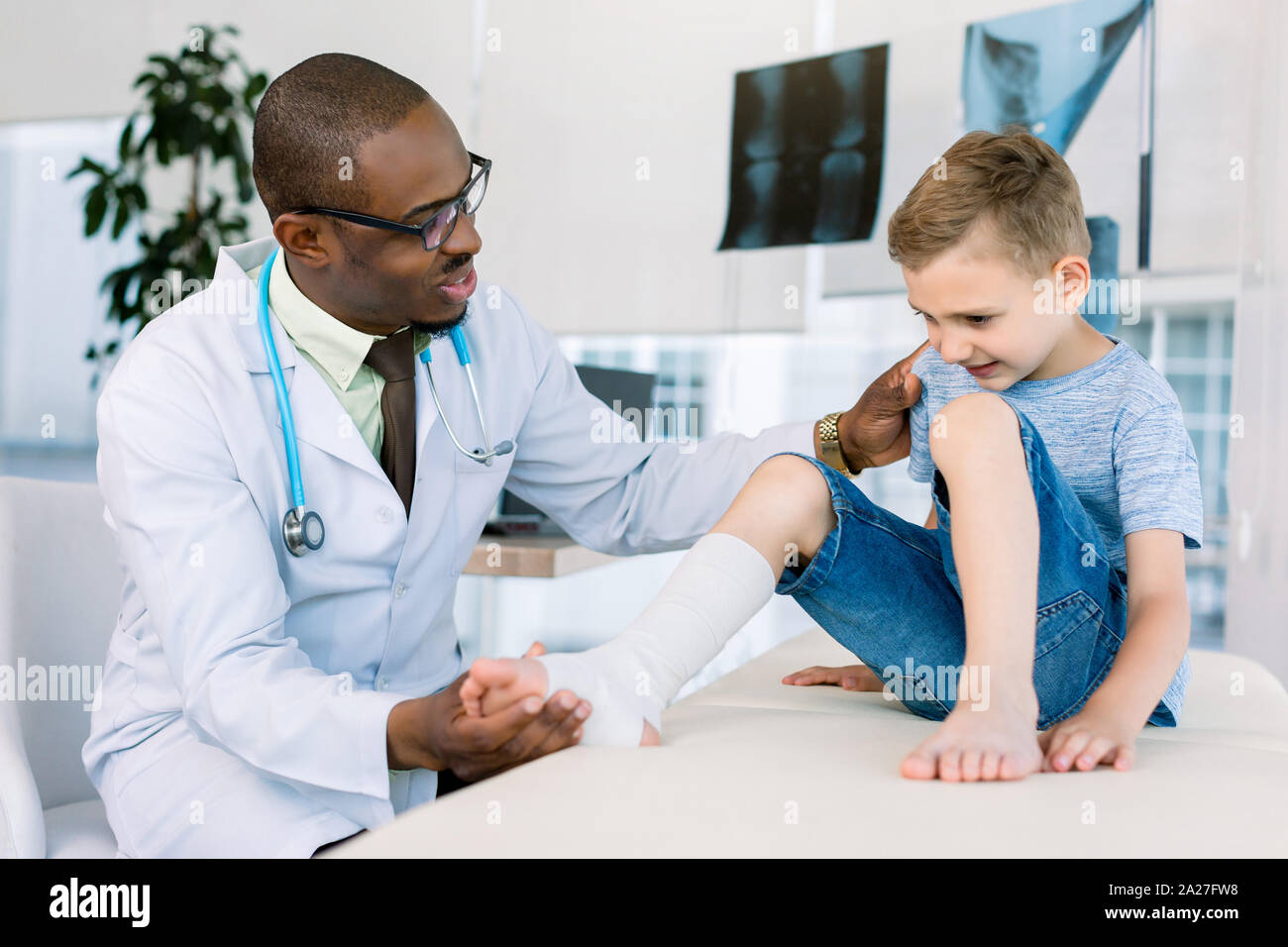 African-American pediatrician examining boy in clinic with leg sprain ...