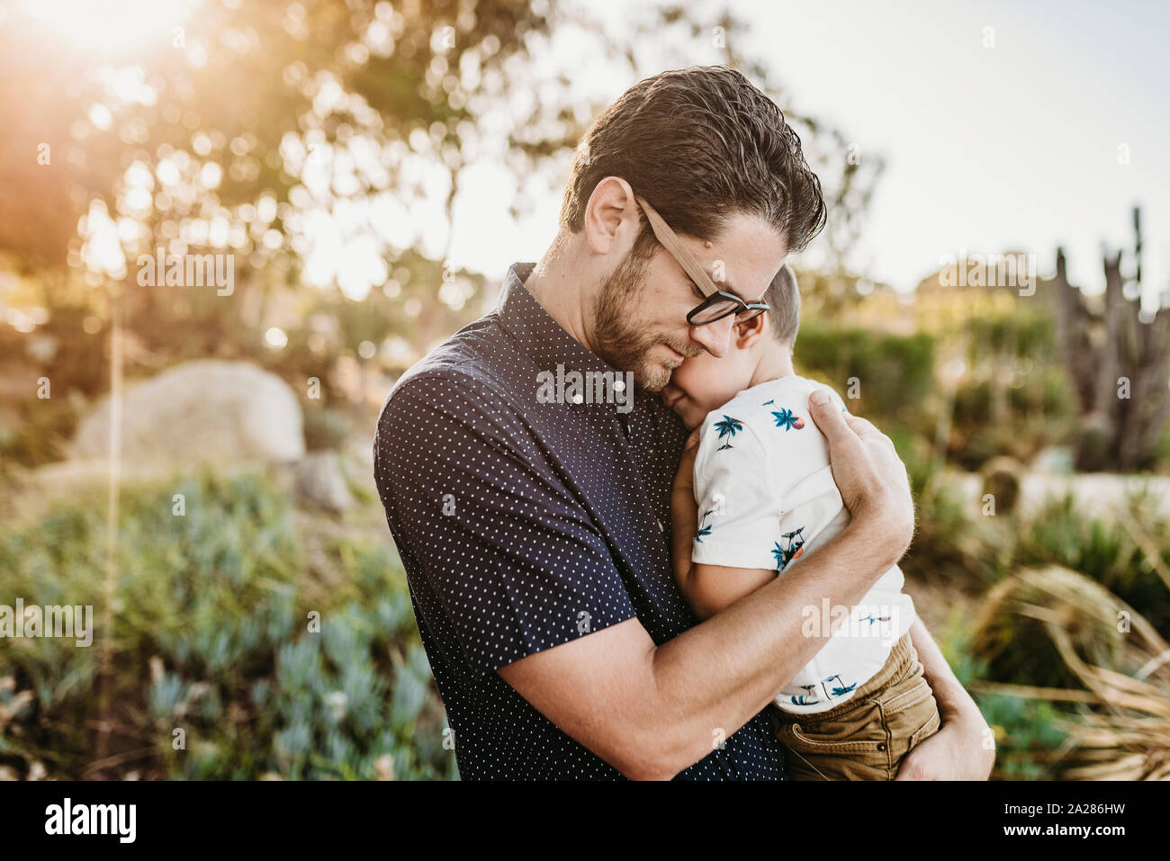 Portrait of father embracing young toddler son with sun behind them ...
