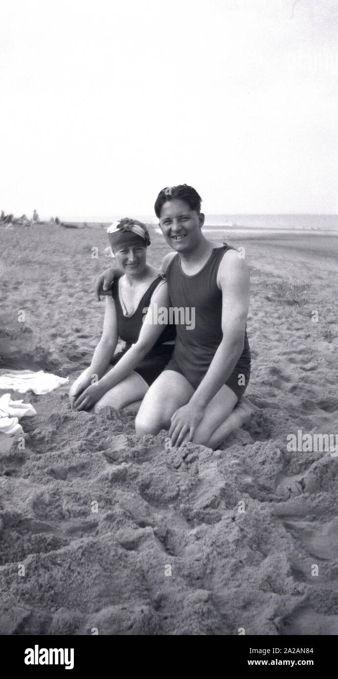 1930s, historical, a man and a lady sitting together on a sandy beach ...