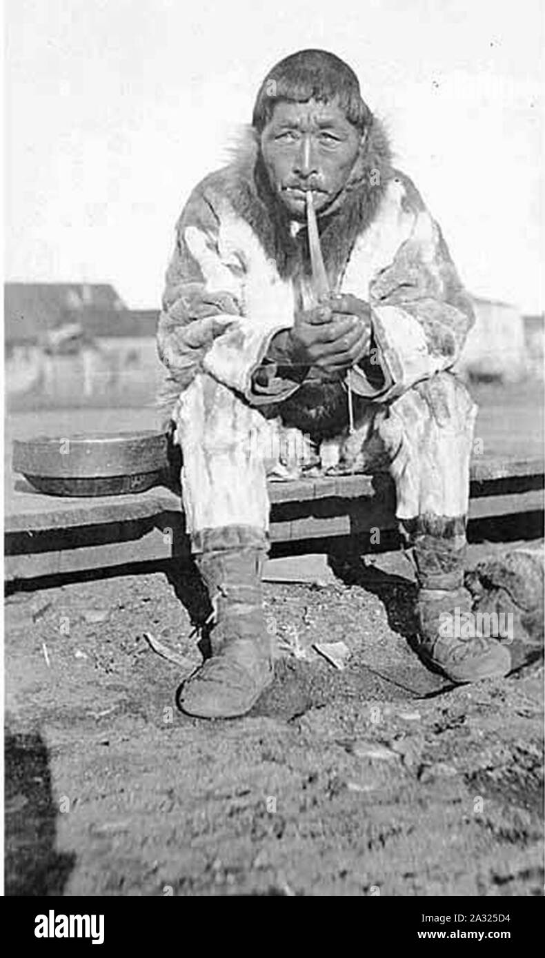 Eskimo man seated on boardwalk smoking pipe wooden dish beside him ...