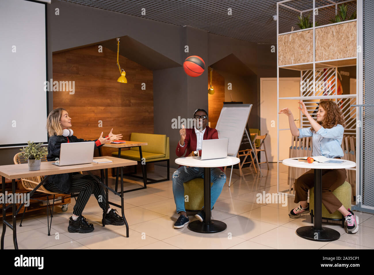 Three happy young students throwing ball to each other while sitting in ...
