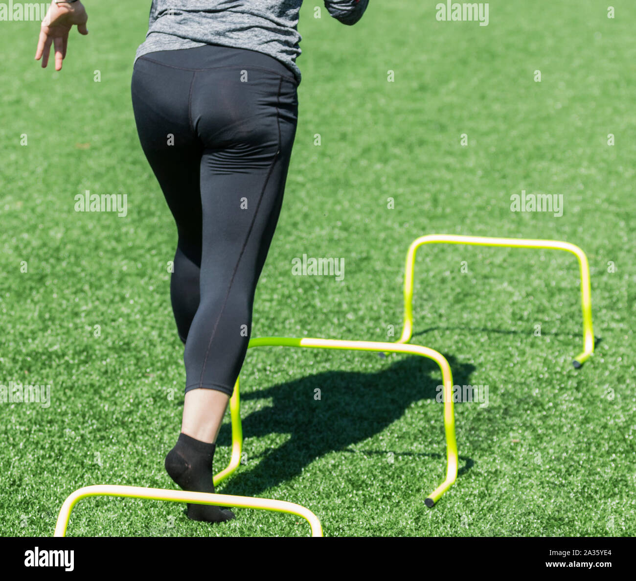 A female athlete is running over yellow mini hurdles on a green turf field during speed and ...