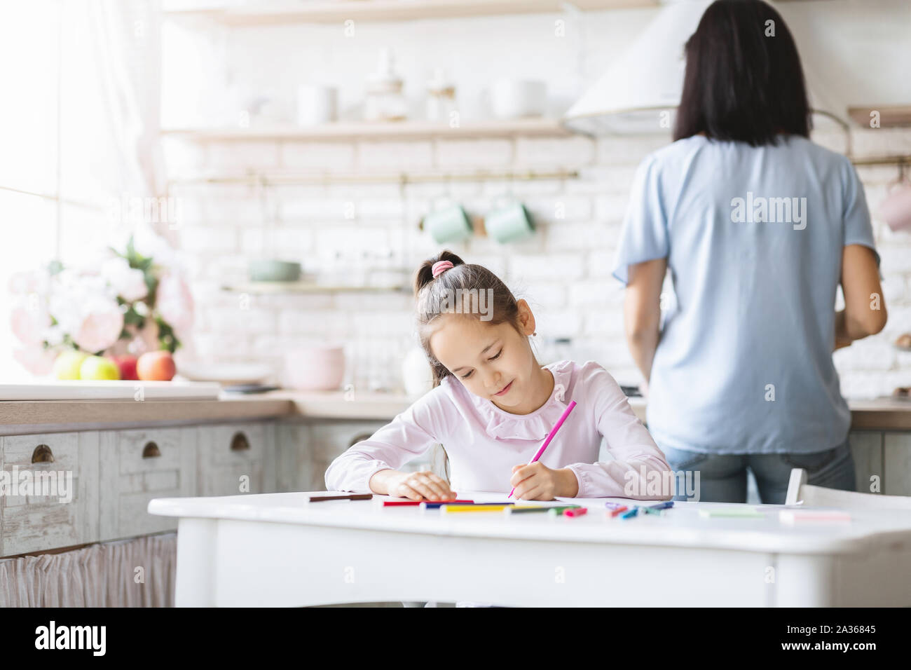 Schoolgirl doing homework in kitchen while her mother cooking food ...