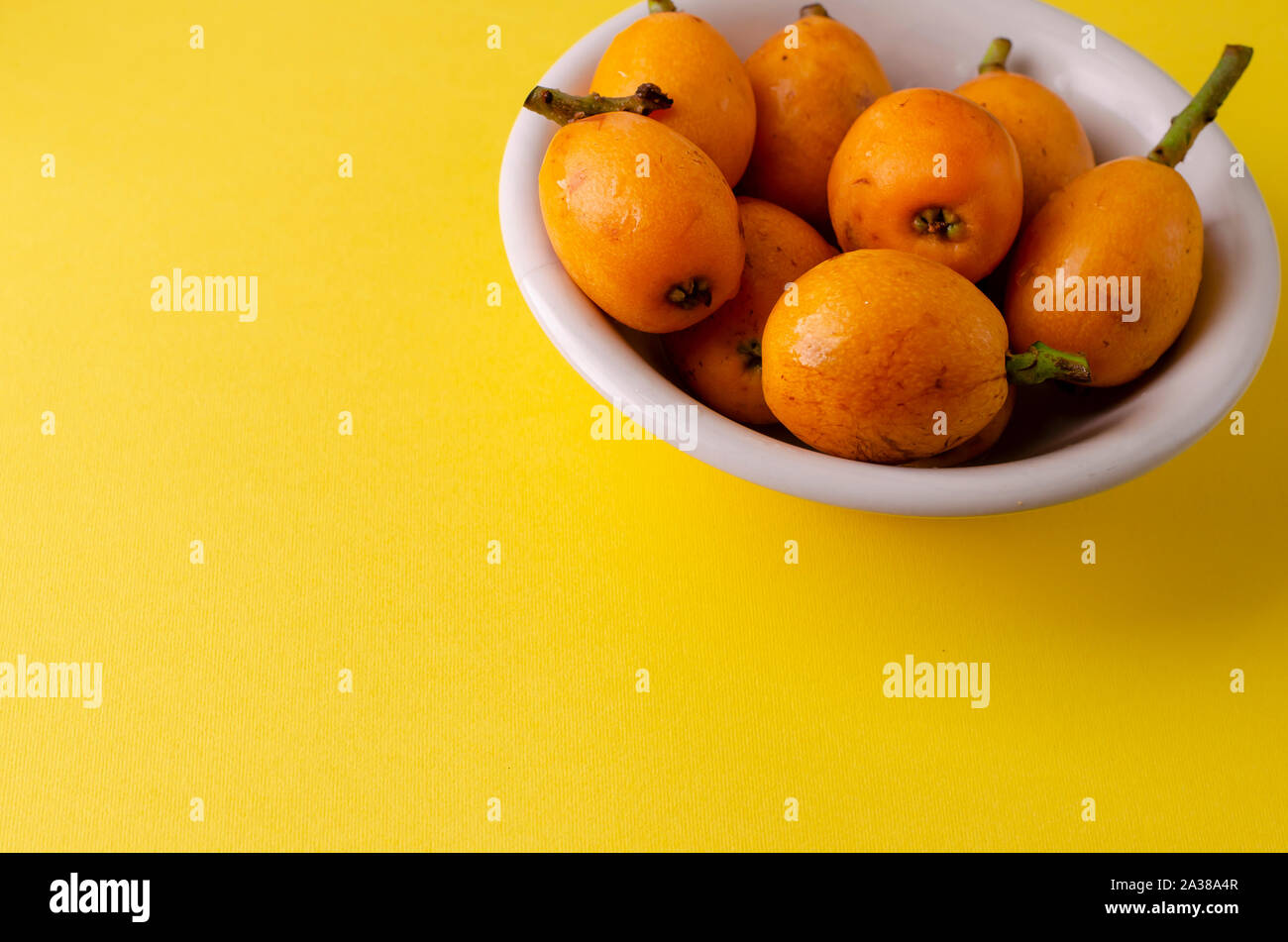 Loquat fruits in a white bowl on bright yellow background with a space ...
