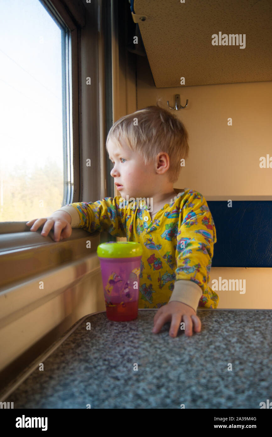 A bright, cute three-year-old boy riding a train looks out the window ...