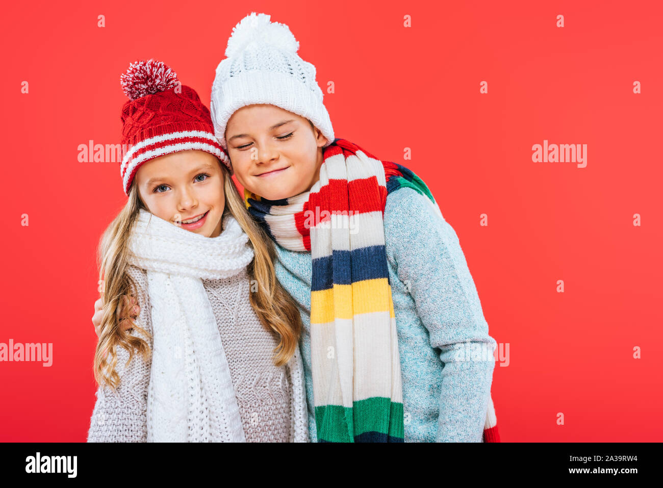 two smiling kids in hats and scarfs embracing isolated on red Stock ...