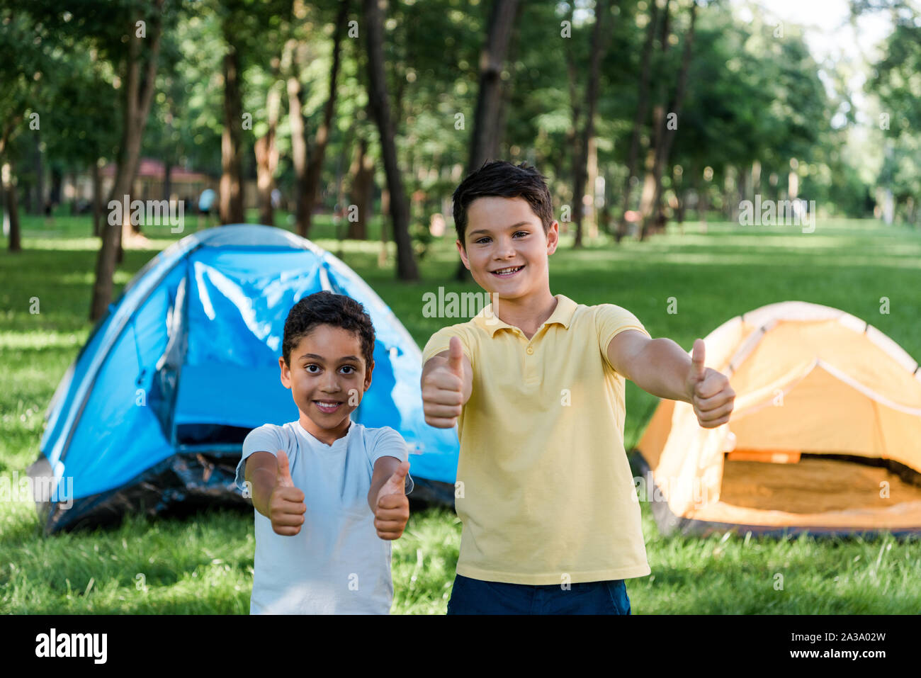 happy multicultural boys smiling while showing thumbs up near camps ...