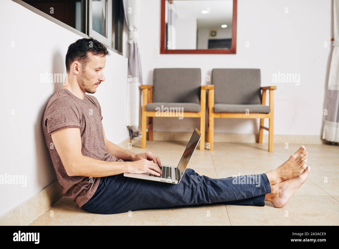 Barefoot young man sitting on the floor under opened window and ...