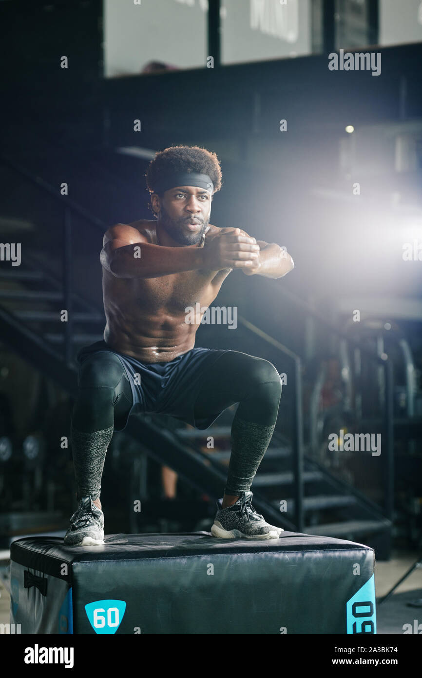 Concentrated muscular young man breathing out when jumping on gym cube ...