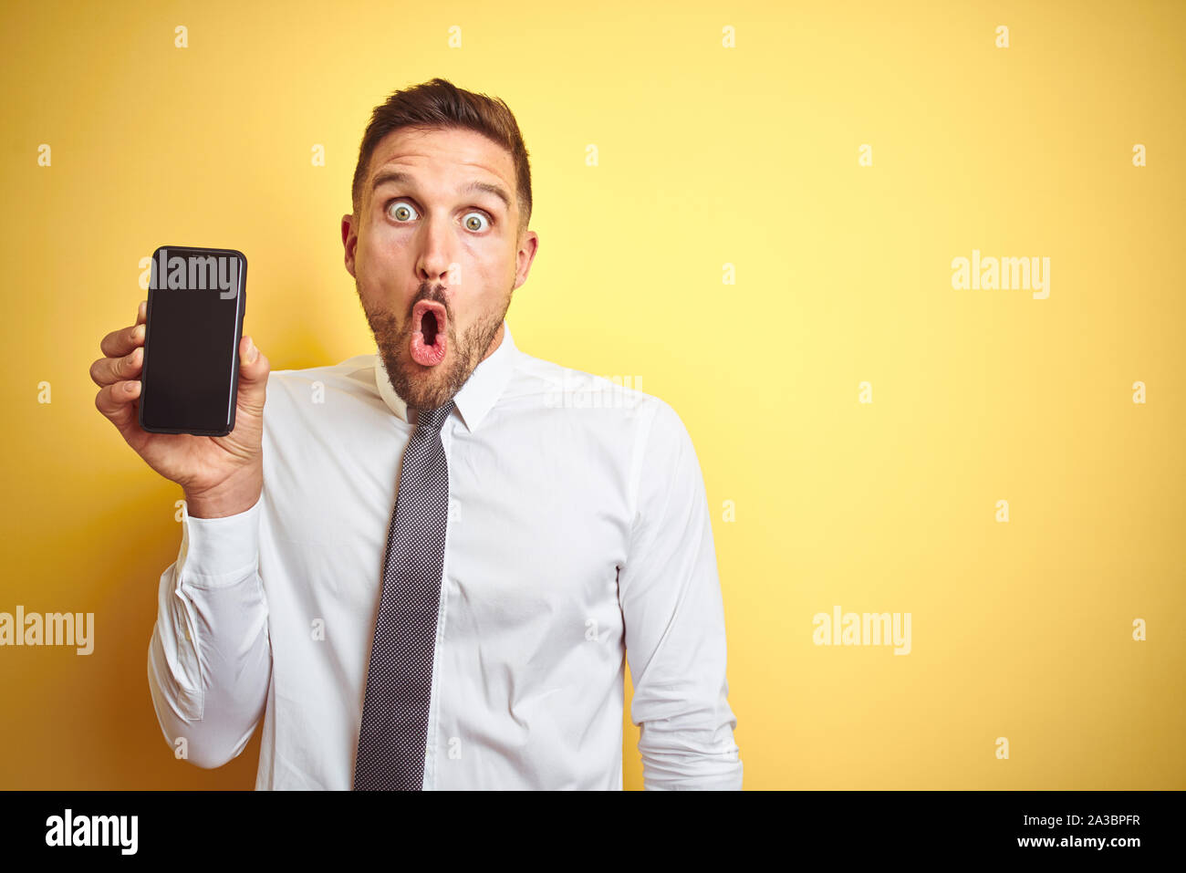 Young handsome business man showing smartphone screen over yellow isolated background scared in ...