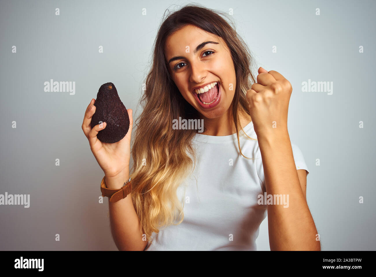 Young beautiful woman eating avocado over grey isolated background ...