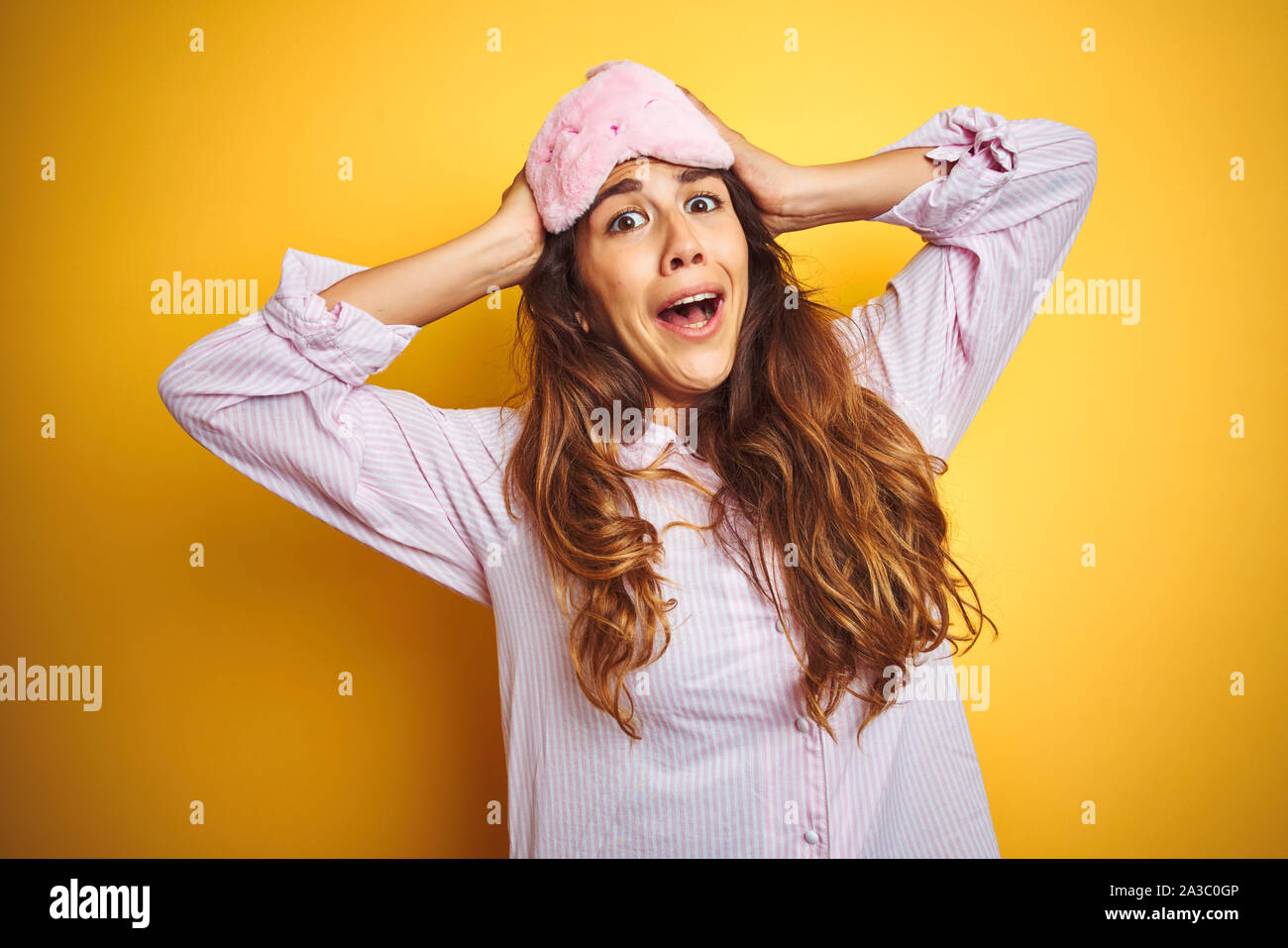 Young woman wearing pajama and sleep mask standing over yellow isolated ...