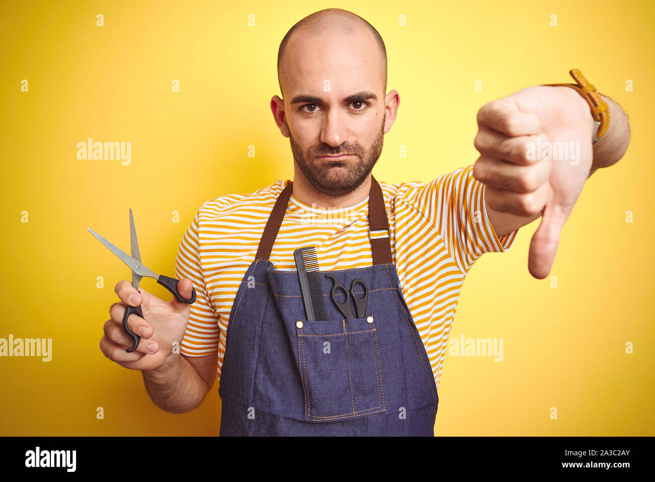 Young hairdresser man wearing barber apron holding scissors over isolated yellow background with ...