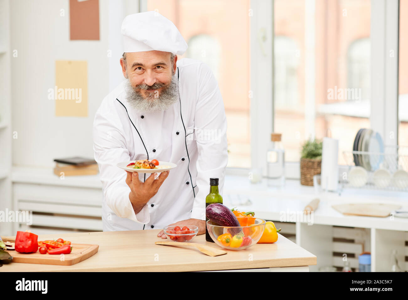 Portrait of cheerful senior chef presenting beautiful dish while posing in restaurant kitchen ...