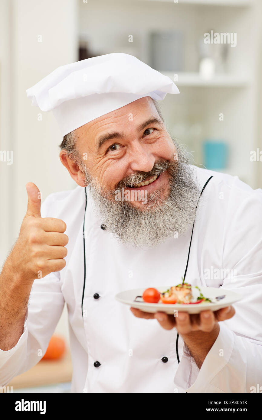 Portrait of cheerful senior chef presenting beautiful Italian dish and ...