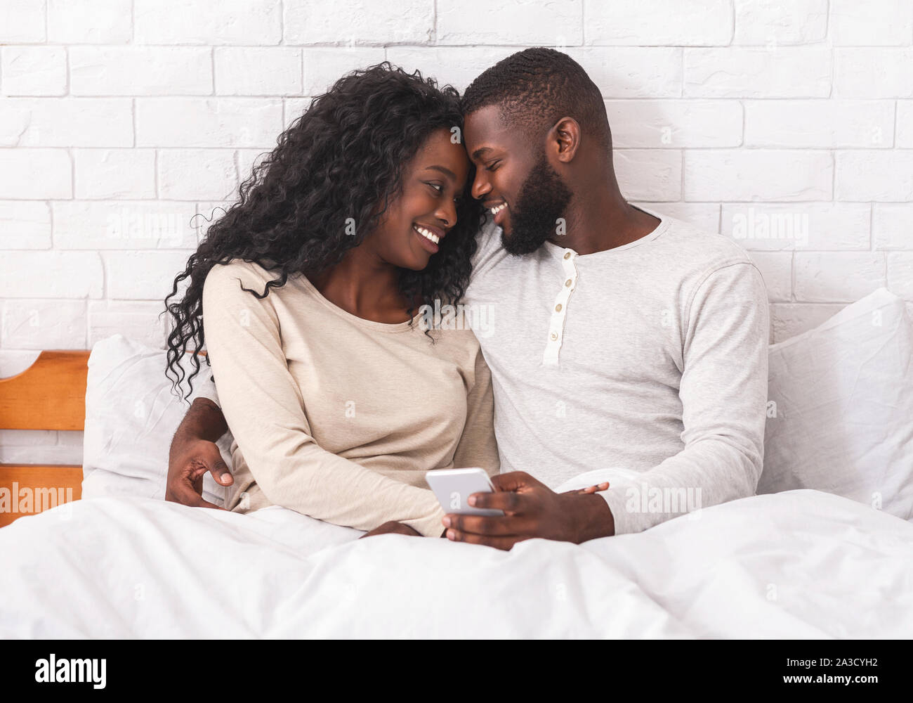 Couple cuddling in bed touching foreheads and using smartphone together ...