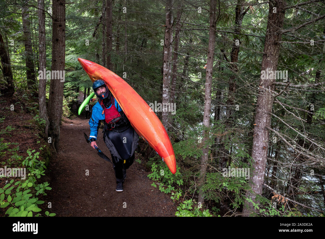 A kayaker walks up a trail from the river carrying the kayak on his ...