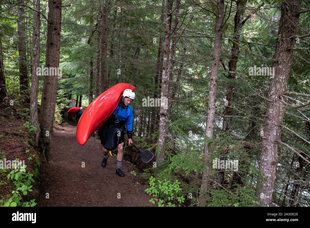 A kayaker walks up a trail from the river carrying the kayak on his ...