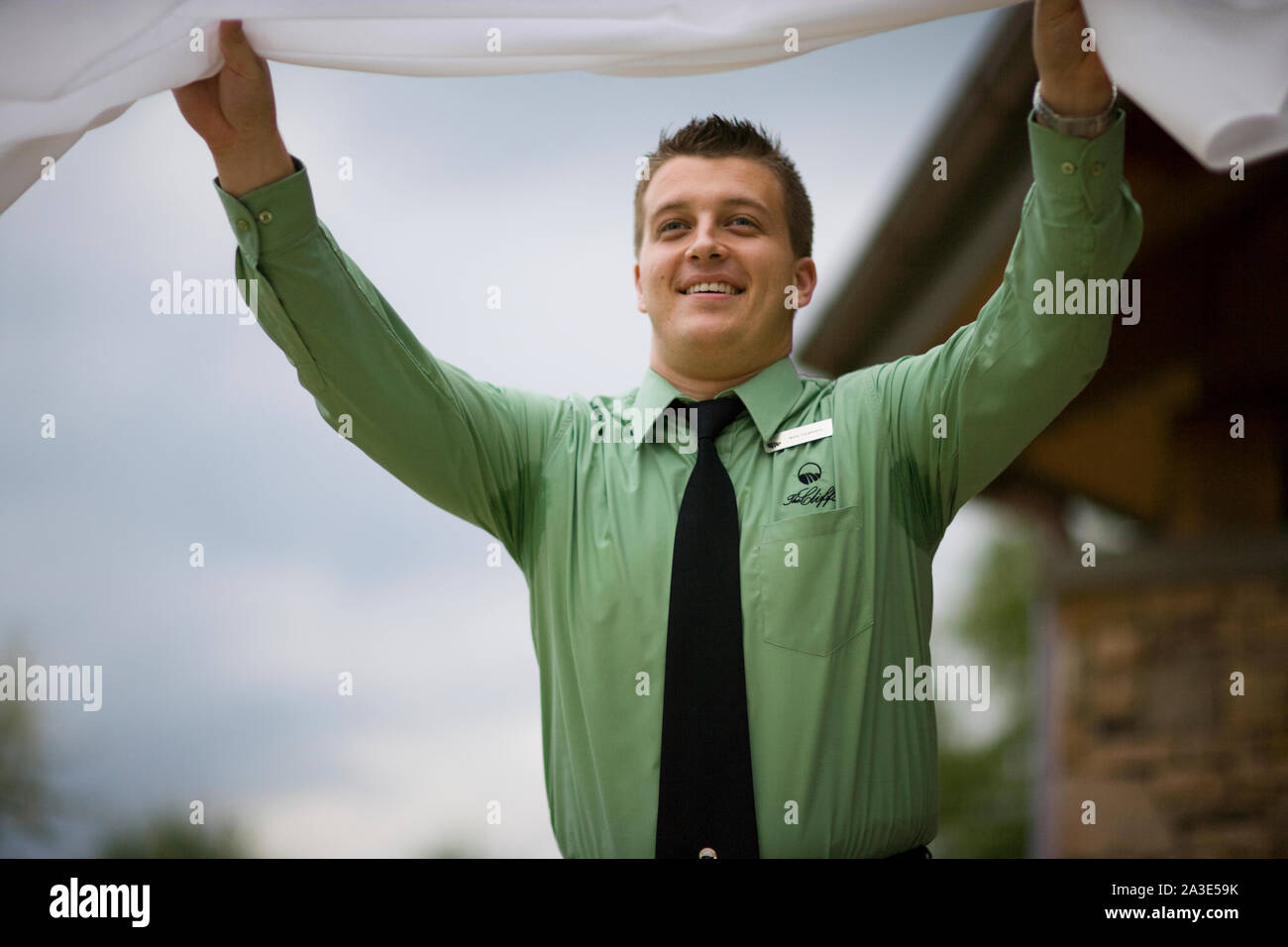 Young adult waiter laying a fresh table cloth at an outside table of a ...