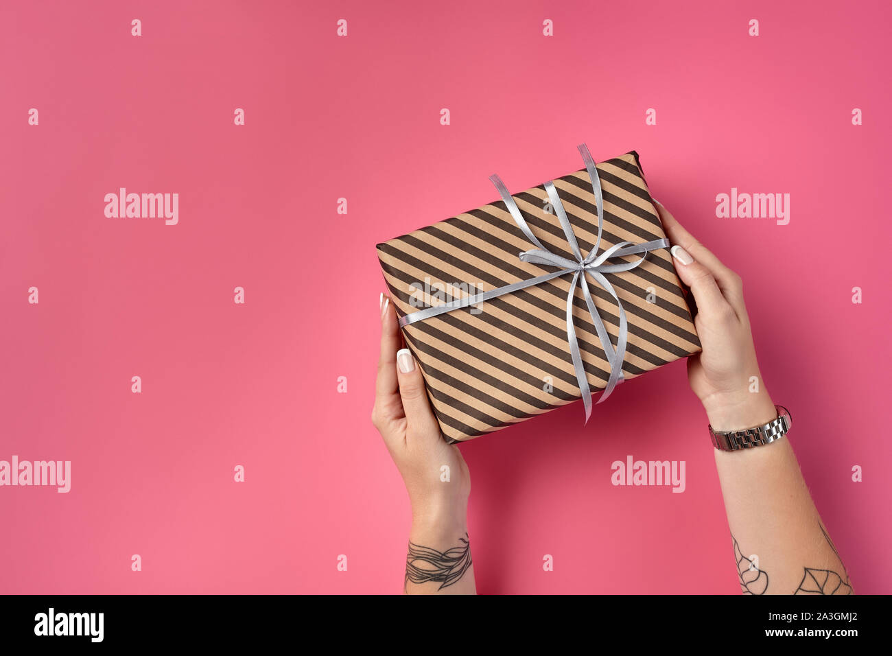 Hands of a woman with tattoos and watch holding striped brown gift box tied with silver ribbon ...