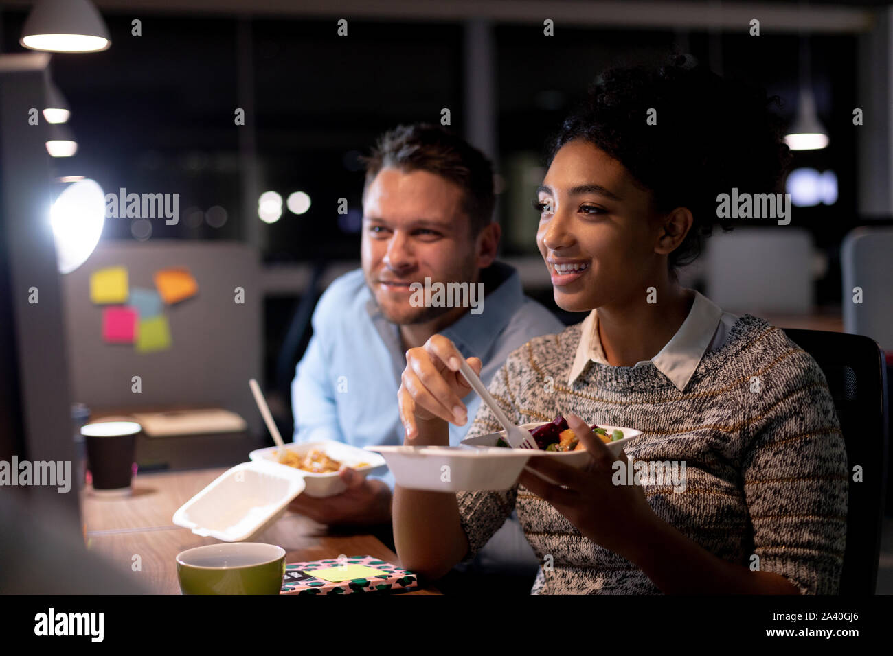 Two young creative professionals working late in a modern office eating ...